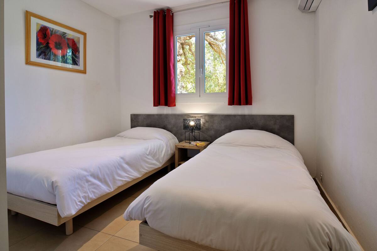 Quarto com duas camas de solteiro, uma mesa de cabeceira com abajur, um quadro na parede e uma janela com cortinas vermelhas.