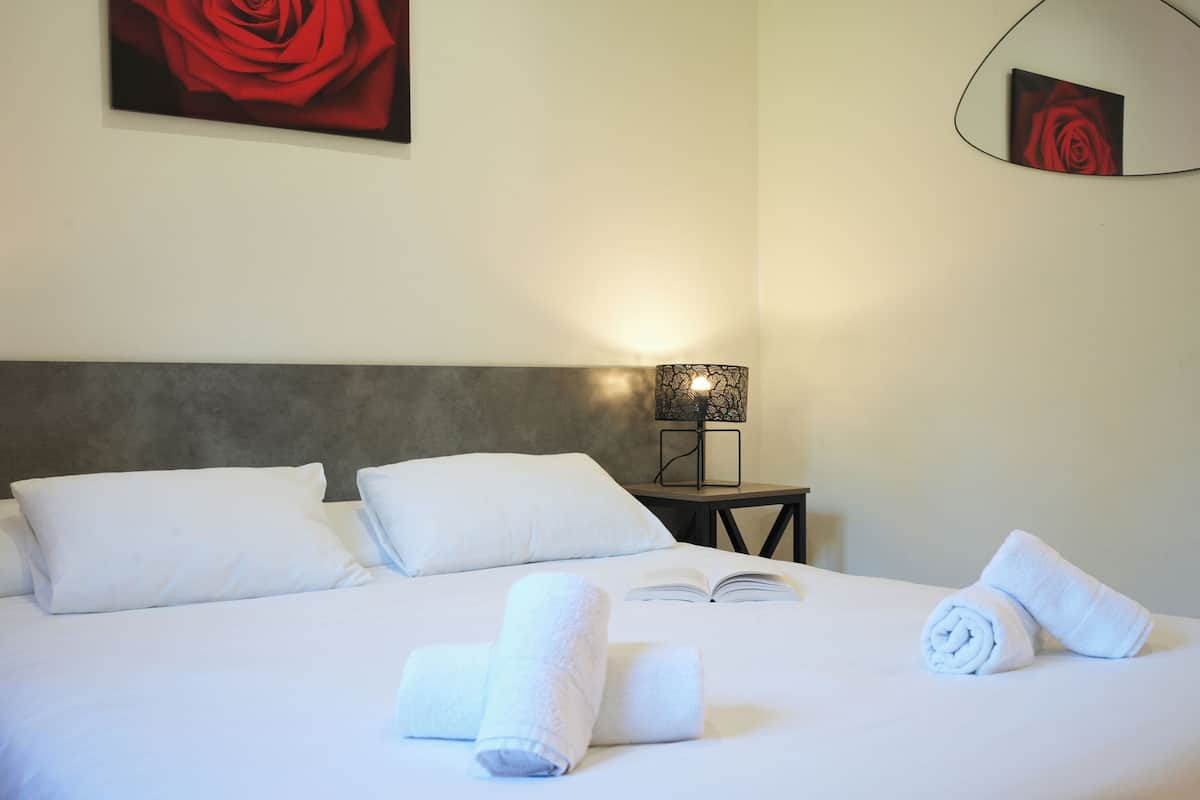 Cama bem arrumada com lençóis brancos, uma pequena mesa de cabeceira com abajur e duas fotos emolduradas de uma rosa vermelha na parede.