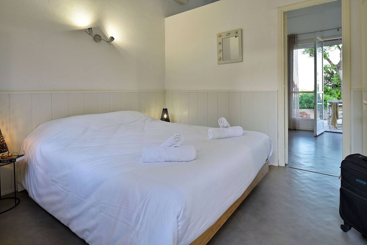 Uma cama grande com lençóis brancos, duas toalhas enroladas, um abajur e uma mala em quarto de hotel com vista para o exterior.