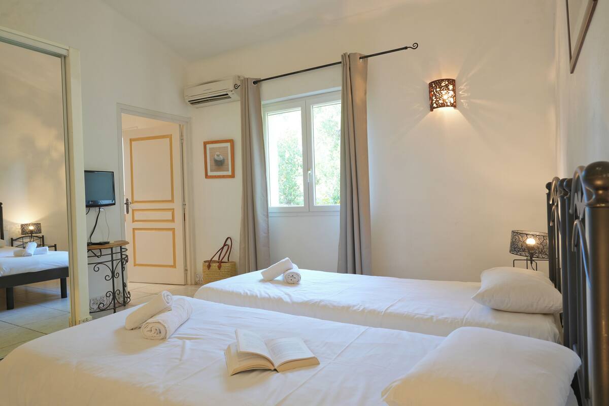 Quarto de hotel com duas camas, televisão, mesa de cabeceira, abajur e janela com cortinas.