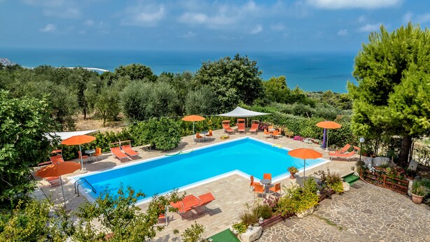 Piscina com espreguiçadeiras e guarda-sóis, cercada por vegetação e vista para o mar.