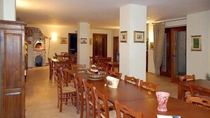 Uma ampla sala de jantar com várias mesas e cadeiras de madeira, uma lareira de pedra e quadros nas paredes.