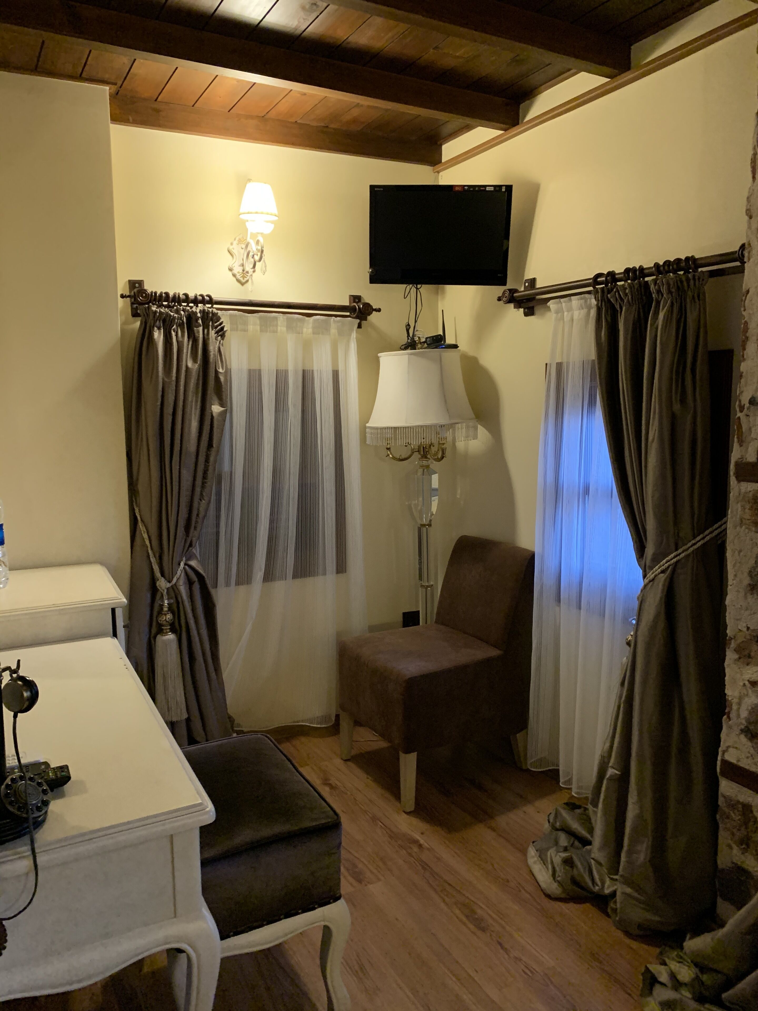 Quarto com teto de madeira, uma mesa branca com telefone, uma cadeira, um abajur e cortinas.