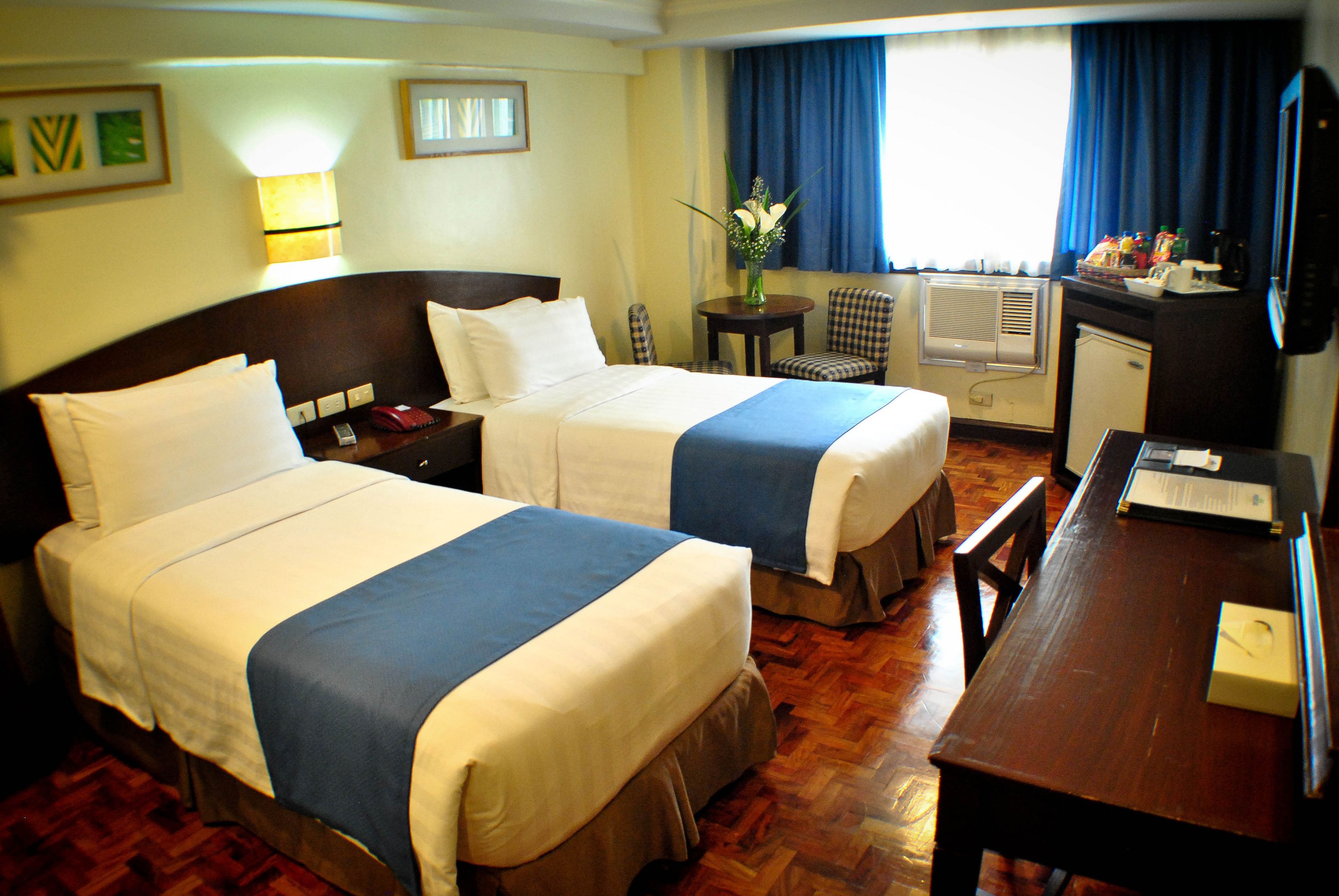 Quarto de hotel com duas camas, uma escrivaninha, uma cadeira, uma mesinha com um vaso de flores e uma televisão.