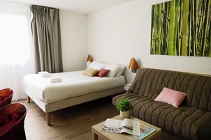 Quarto de hotel com uma cama, um sofá, uma mesinha com uma revista, uma planta e uma parede com estampa de bambu.