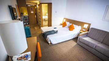 Quarto de hotel com cama, sofá, escrivaninha, televisão e banheiro visível por uma porta aberta.