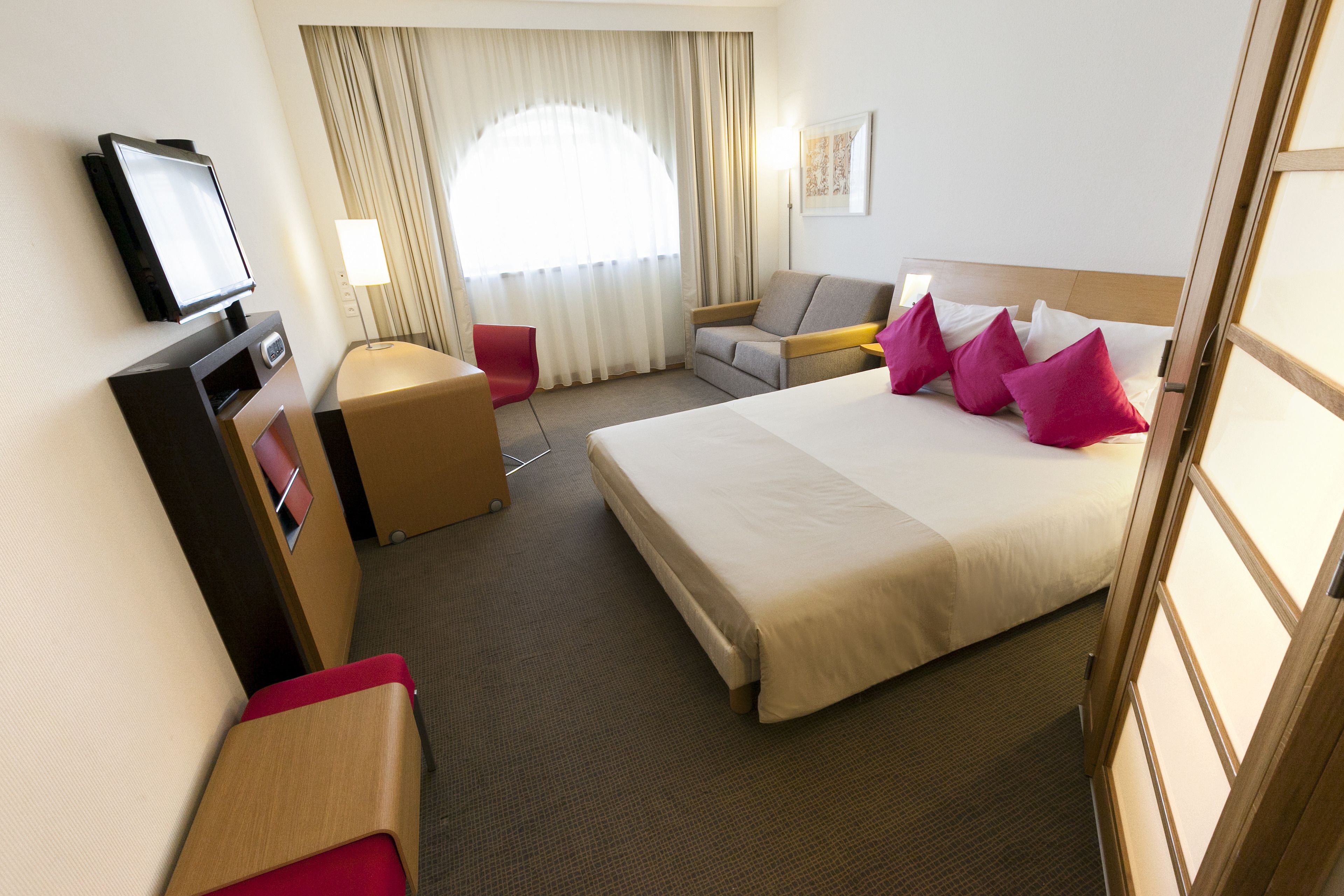 Quarto de hotel com cama, escrivaninha, cadeira, televisão e janela com cortinas.