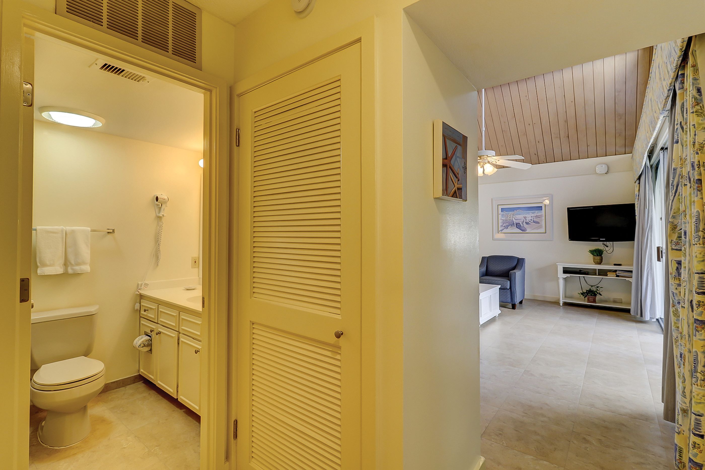 Casa de banho com sanita, lavatório e suporte para toalhas. Uma porta dá para uma área de estar com televisão, uma cadeira e uma planta.