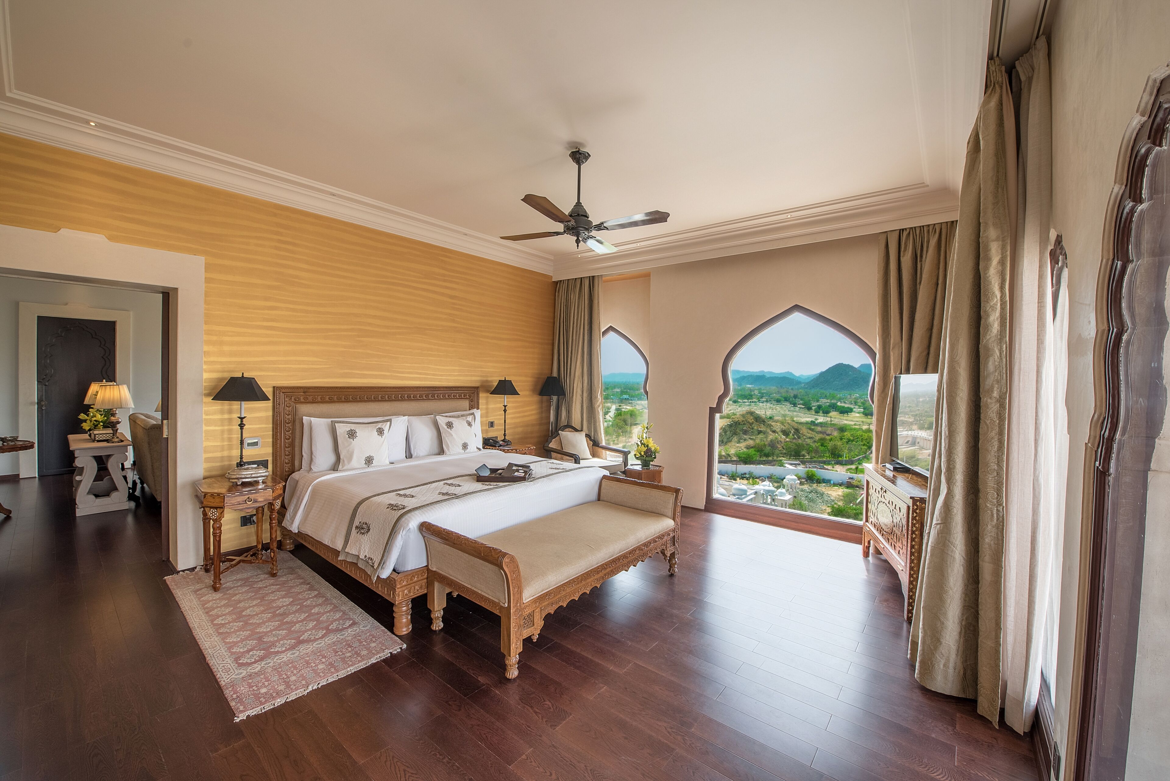 Um quarto espaçoso com uma cama grande, um ventilador de teto e vista para uma paisagem montanhosa através de janelas em arco.