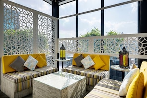Uma área de estar moderna ao ar livre, com sofás listrados e uma mesa de centro de vidro, cercada por painéis de treliça decorativos e duas lanternas.