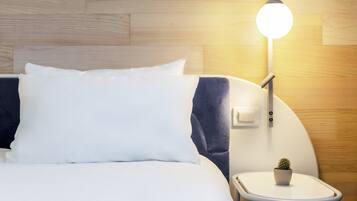Cama bem arrumada com roupa de cama branca e azul marinho, mesa de cabeceira com um pequeno vaso de planta e luminária fixada na parede.