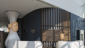 Uma área de recepção de hotel moderna, com um balcão curvo, duas grandes luminárias de vime suspensas e uma parede escura com ripas verticais de madeira.