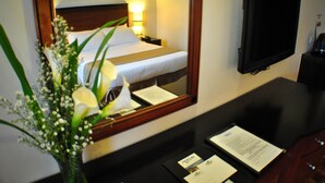 Quarto de hotel com cama, mesa de cabeceira, espelho, televisão e um arranjo floral.