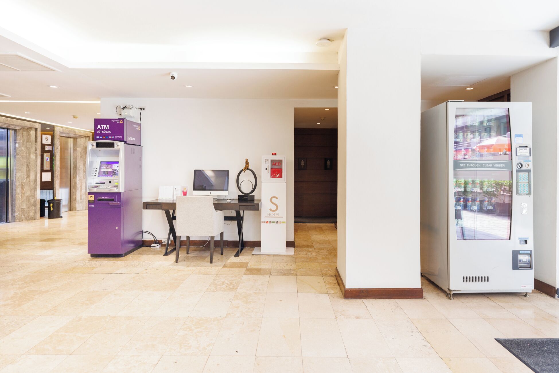 Um saguão de hotel com um caixa eletrônico, um computador, uma mesa de recepção e uma máquina de vendas automáticas.