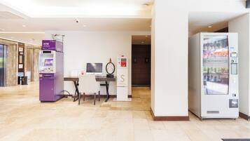 Um saguão de hotel com um caixa eletrônico, um computador, uma mesa de recepção e uma máquina de vendas automáticas.