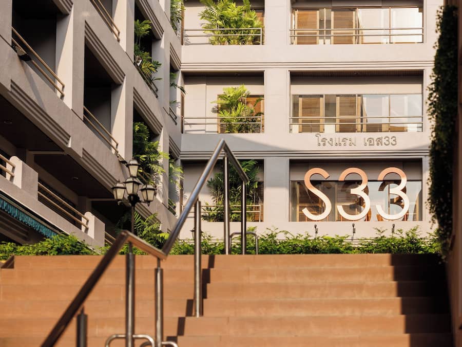 Um edifício com vários andares e uma escadaria que leva à entrada. O edifício possui varandas com plantas, e a entrada tem uma placa grande com o número 'S33'.