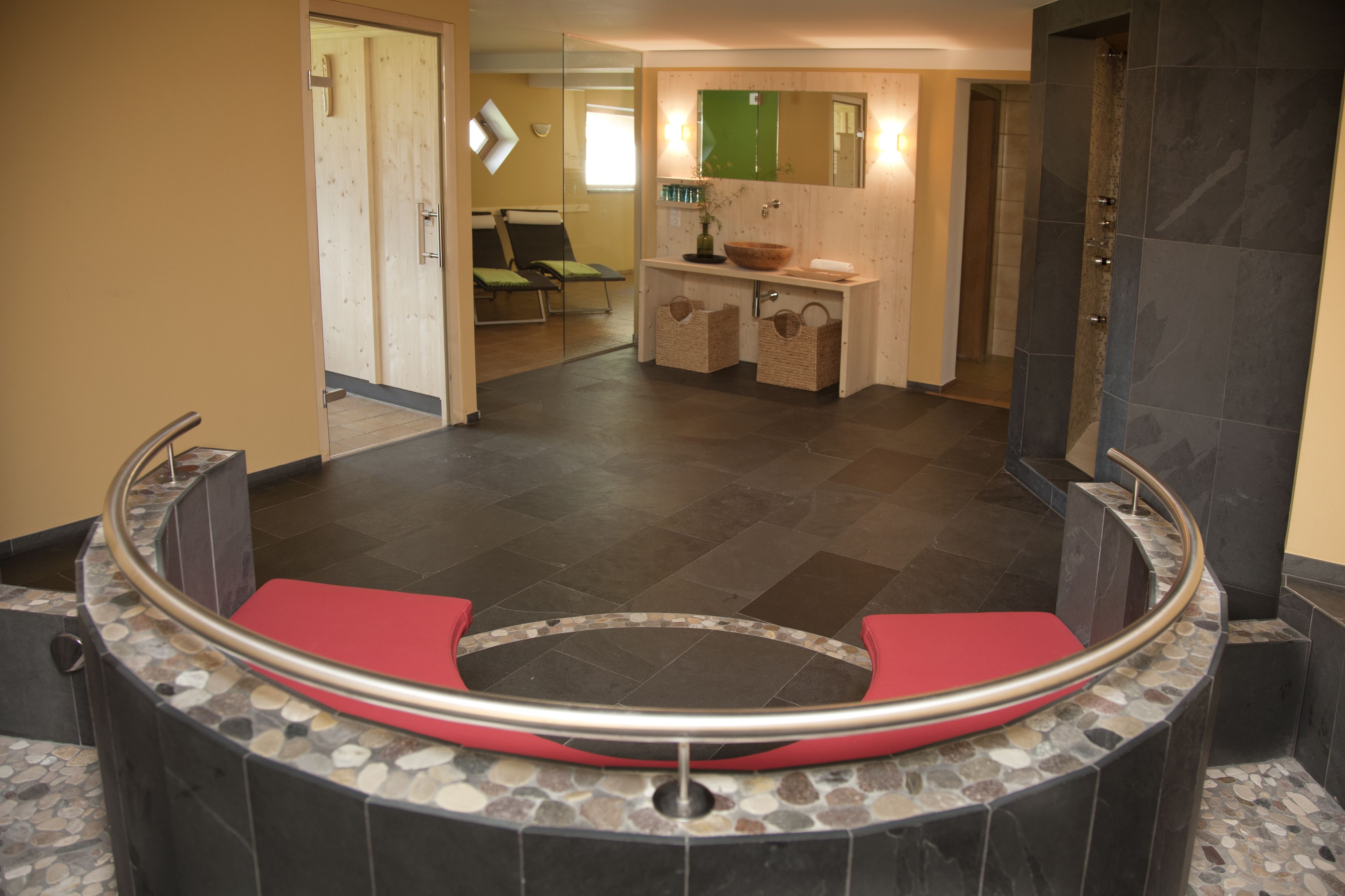 Uma banheira de hidromassagem circular com um assento vermelho, piso de ladrilho de pedra e uma porta de madeira que dá para um quarto com parede verde e um espelho.