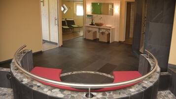 Uma banheira de hidromassagem circular com um assento vermelho, piso de ladrilho de pedra e uma porta de madeira que dá para um quarto com parede verde e um espelho.
