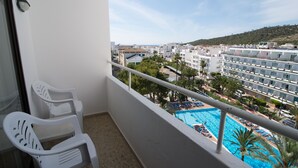 Uma varanda com cadeiras de plástico branco, área de piscina com espreguiçadeiras e vista para um complexo hoteleiro.