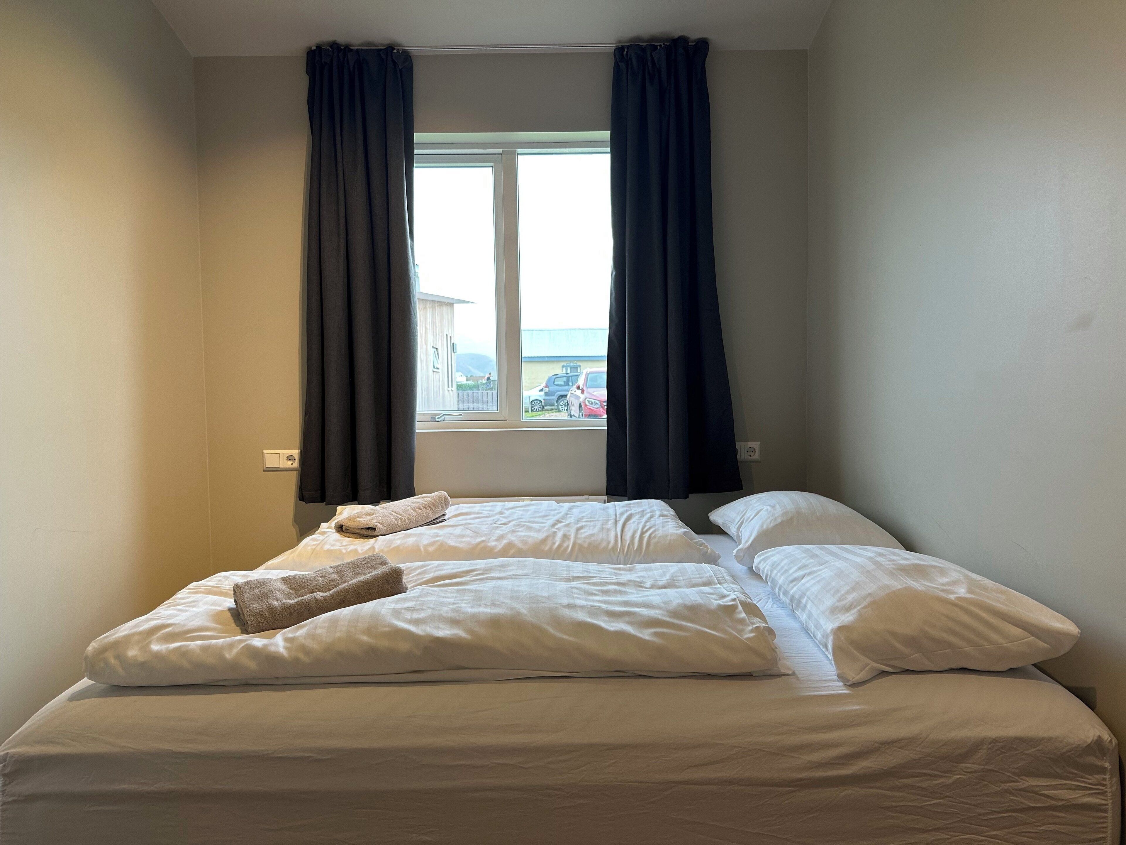 Uma cama de solteiro com lençóis brancos e duas almofadas, uma janela com cortinas escuras e vista para um edifício e veículos lá fora.