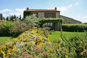 Uma casa de pedra com telhado de telhas vermelhas, rodeada por um jardim bem cuidado com diversas plantas e arbustos floridos.