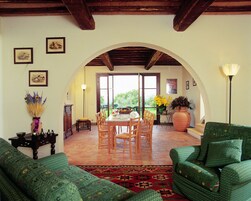 Uma área de refeições com mesa e cadeiras, um vaso grande com flores e vista através de um arco aberto.