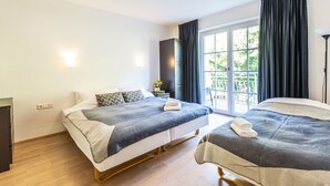 Quarto de hotel com duas camas, piso de madeira, janela com cortinas, mesa de cabeceira com abajur e luminária na parede.