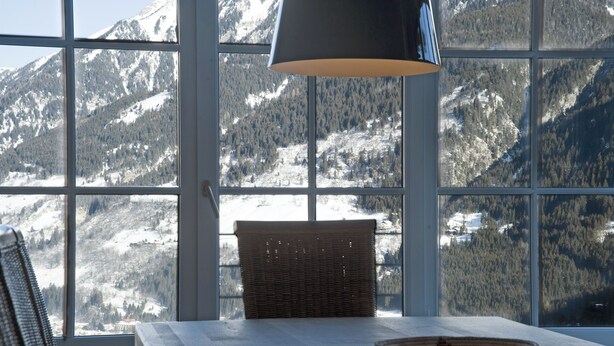 Quarto com uma janela ampla que oferece vista para as montanhas, uma cadeira de vime, uma mesa e um abajur.