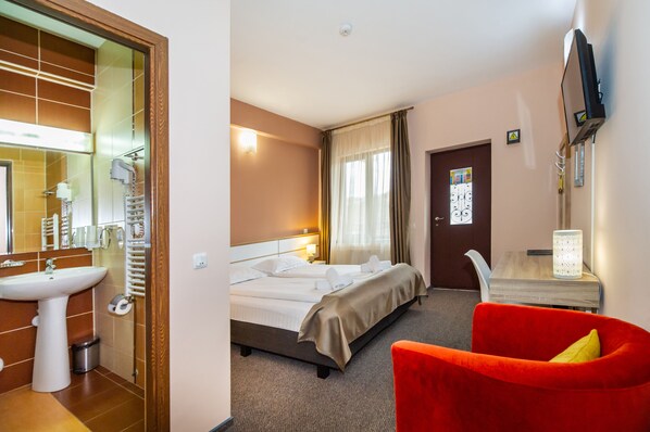 Quarto de hotel com cama, uma cadeira vermelha, mesa de cabeceira com abajur, banheiro com pia e espelho.