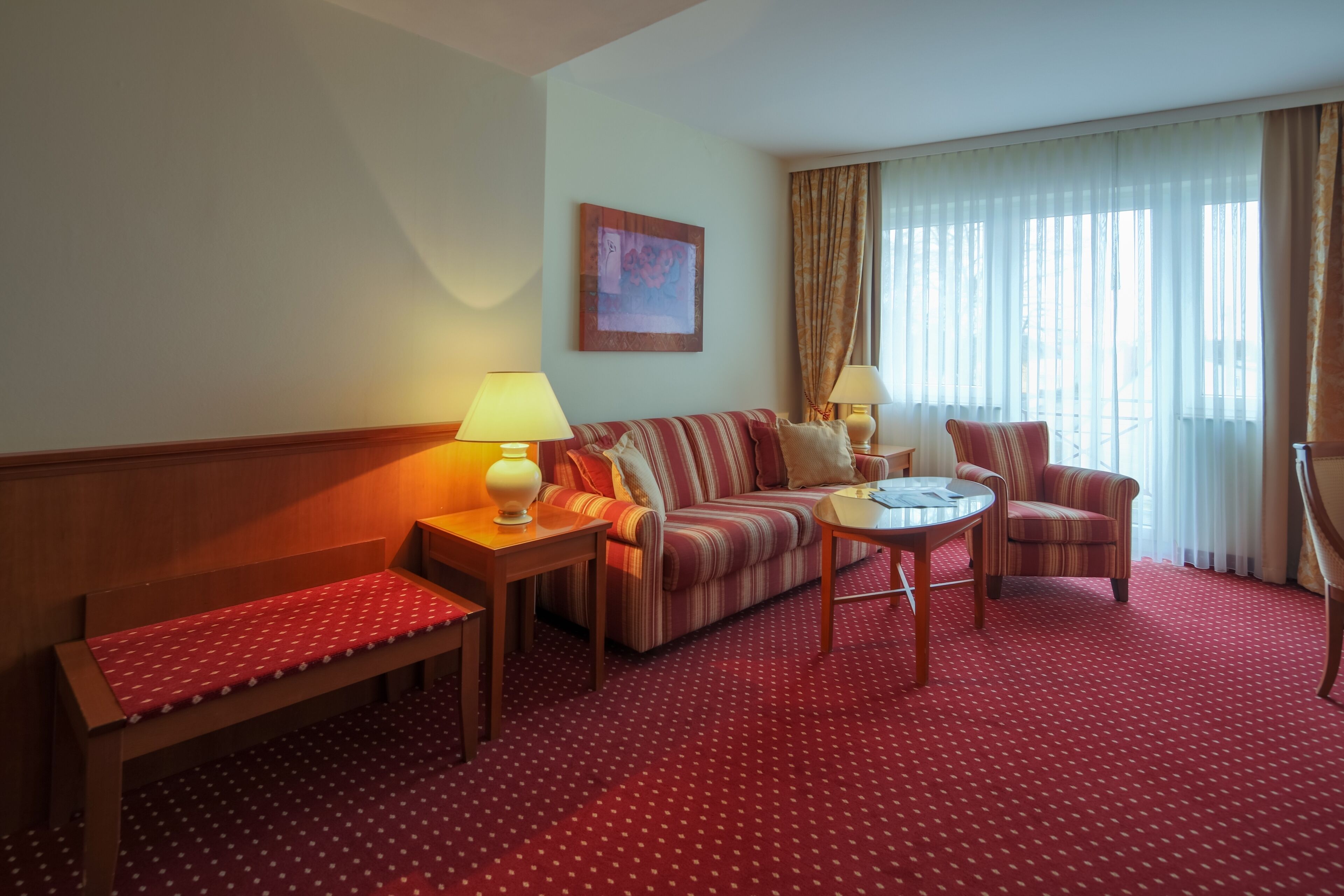 Quarto de hotel com tapete vermelho, sofá, cadeira, mesa pequena e luminária.