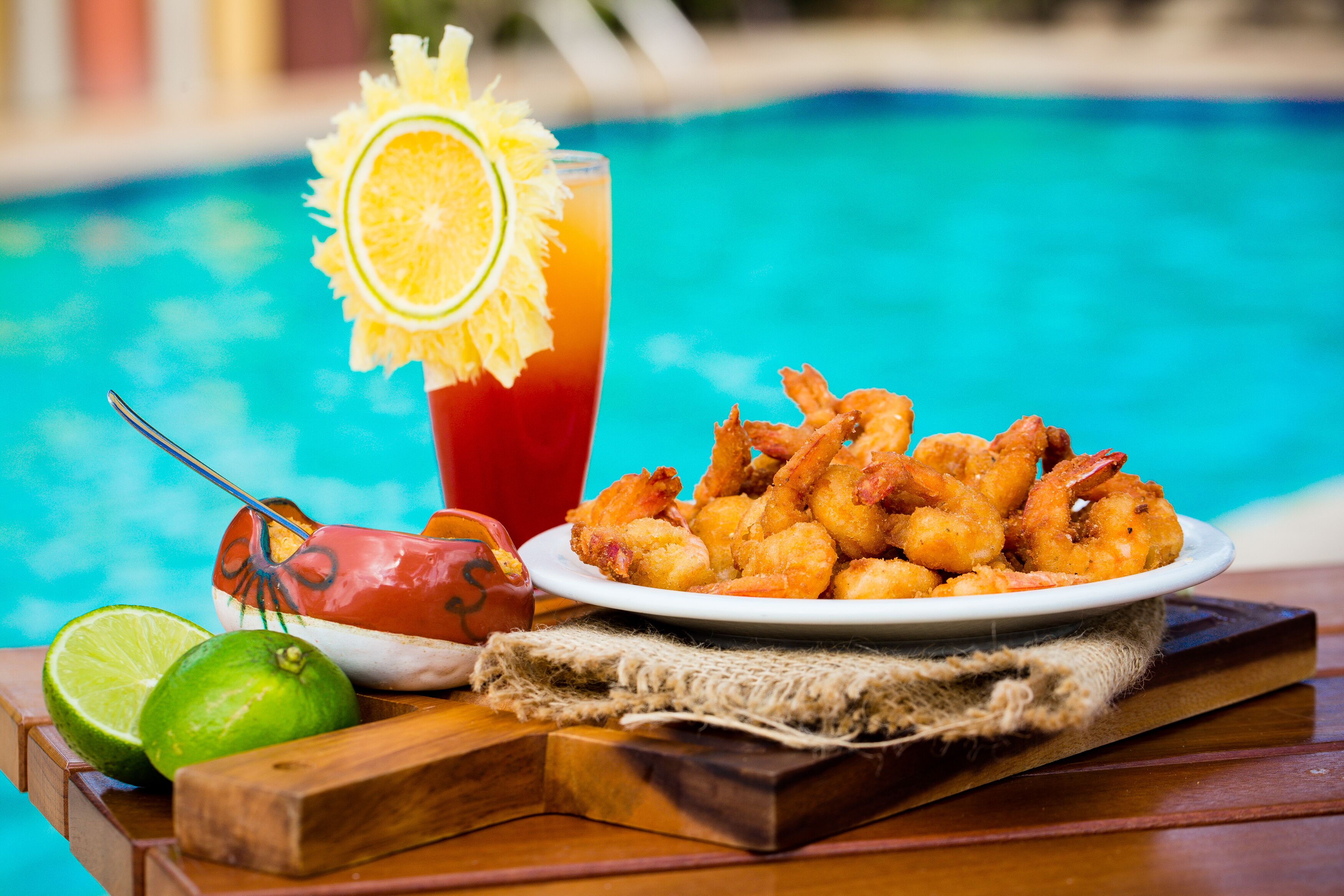 Um prato de camarões fritos e um copo de bebida vermelha com um toque de limão, apoiados sobre uma mesa de madeira junto a uma piscina.
