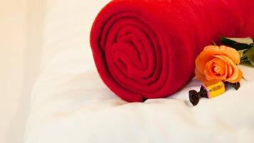 Uma cama bem arrumada com roupa de cama branca e laranja, uma toalha vermelha enrolada e uma única rosa laranja.