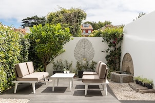 Um pátio externo com dois sofás bege, uma mesa branca e um mural decorativo com folhas na parede.