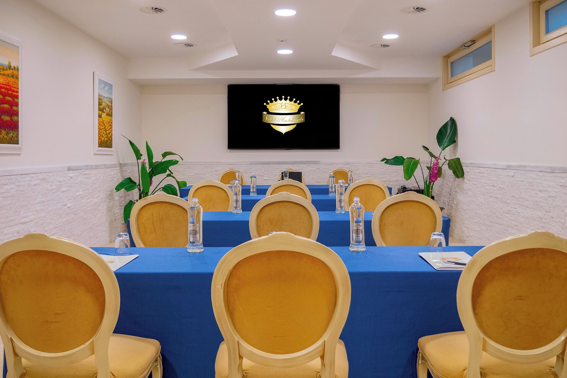 Uma sala de conferências com uma mesa azul, cadeiras amarelas e uma tela exibindo um logotipo de coroa.
