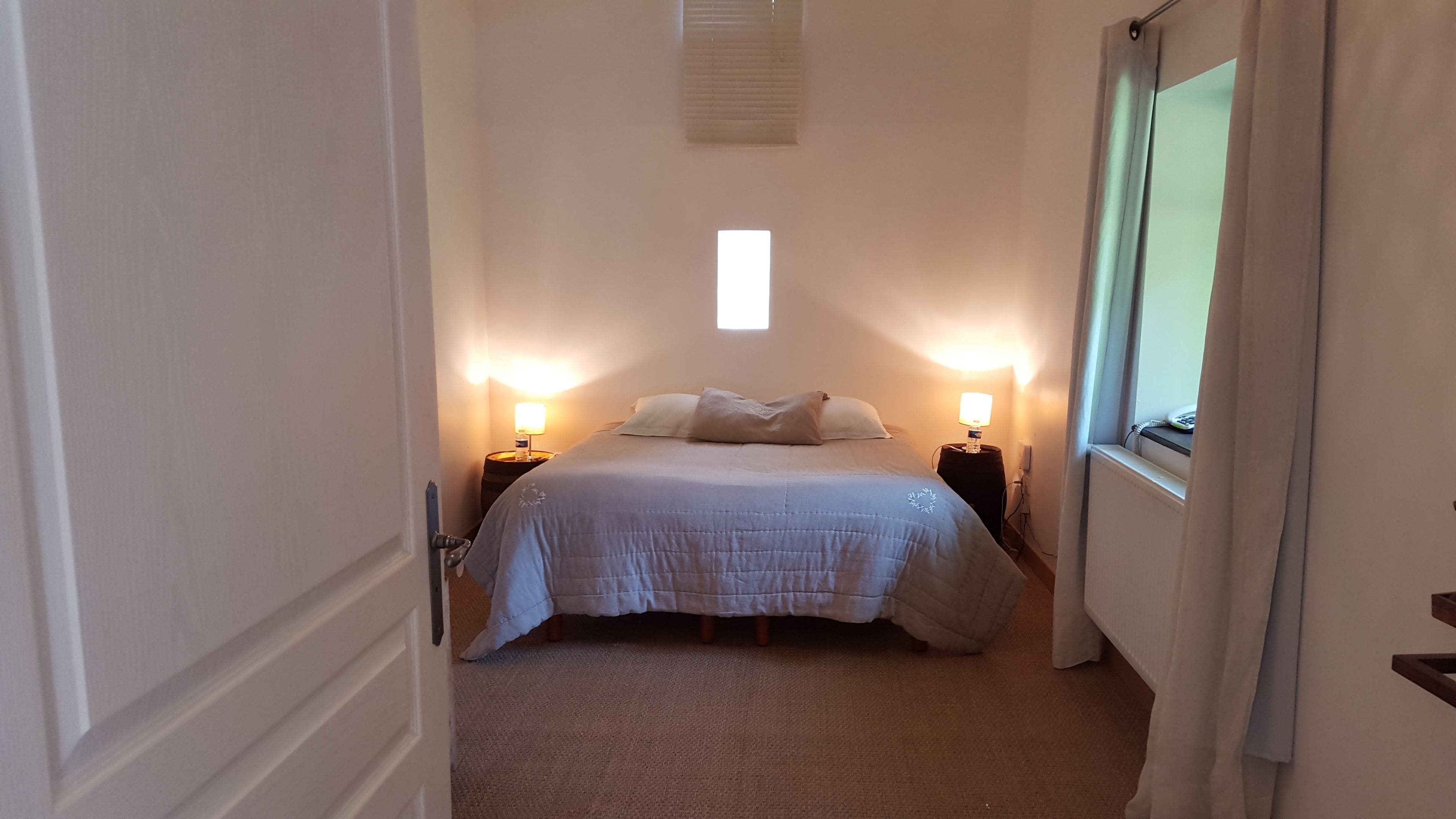 Um corredor estreito que leva a um quarto com uma cama, duas luminárias de cabeceira e uma janela.