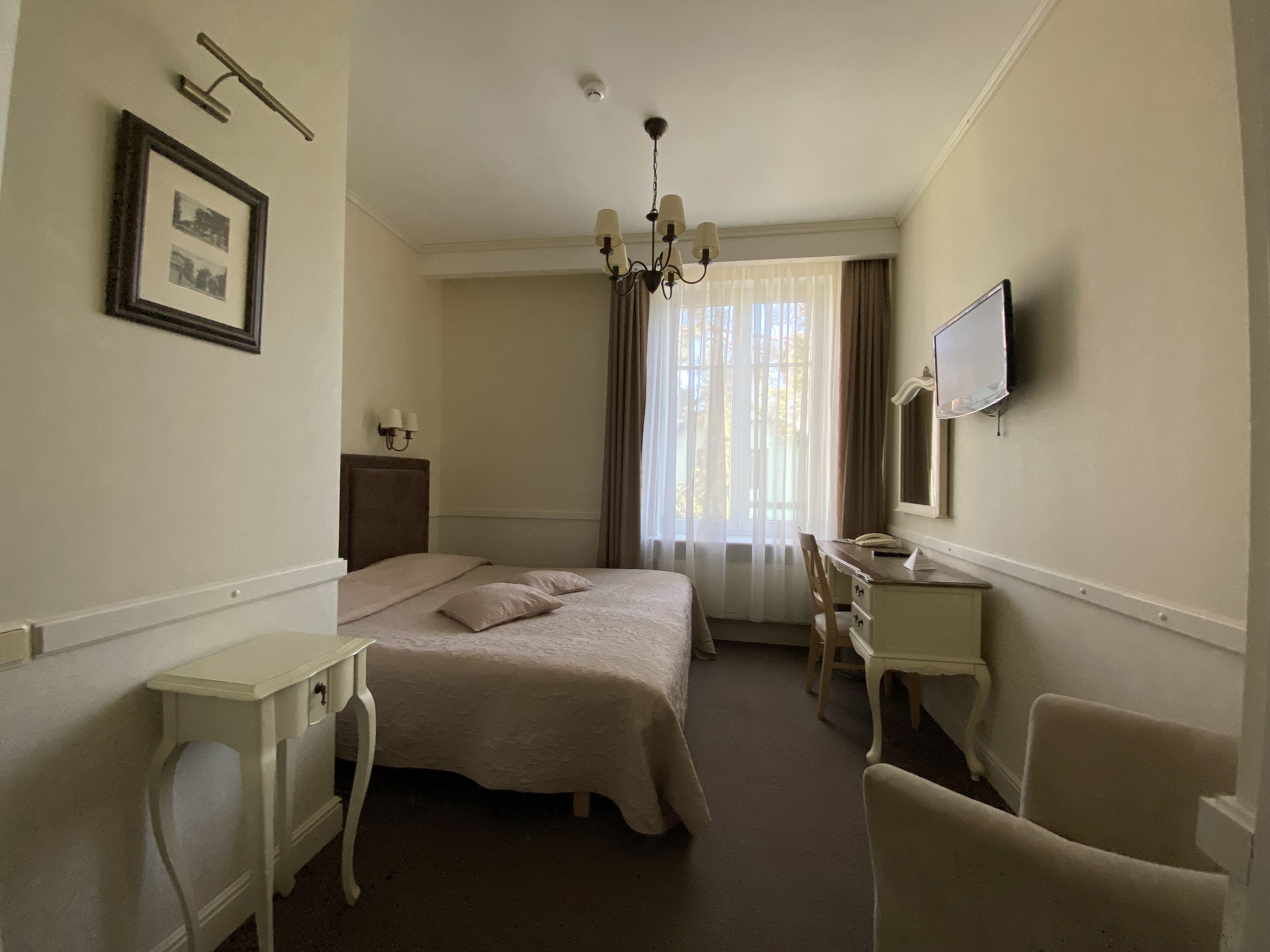 Quarto de hotel com cama, escrivaninha, cadeira, televisão e janela com cortinas.