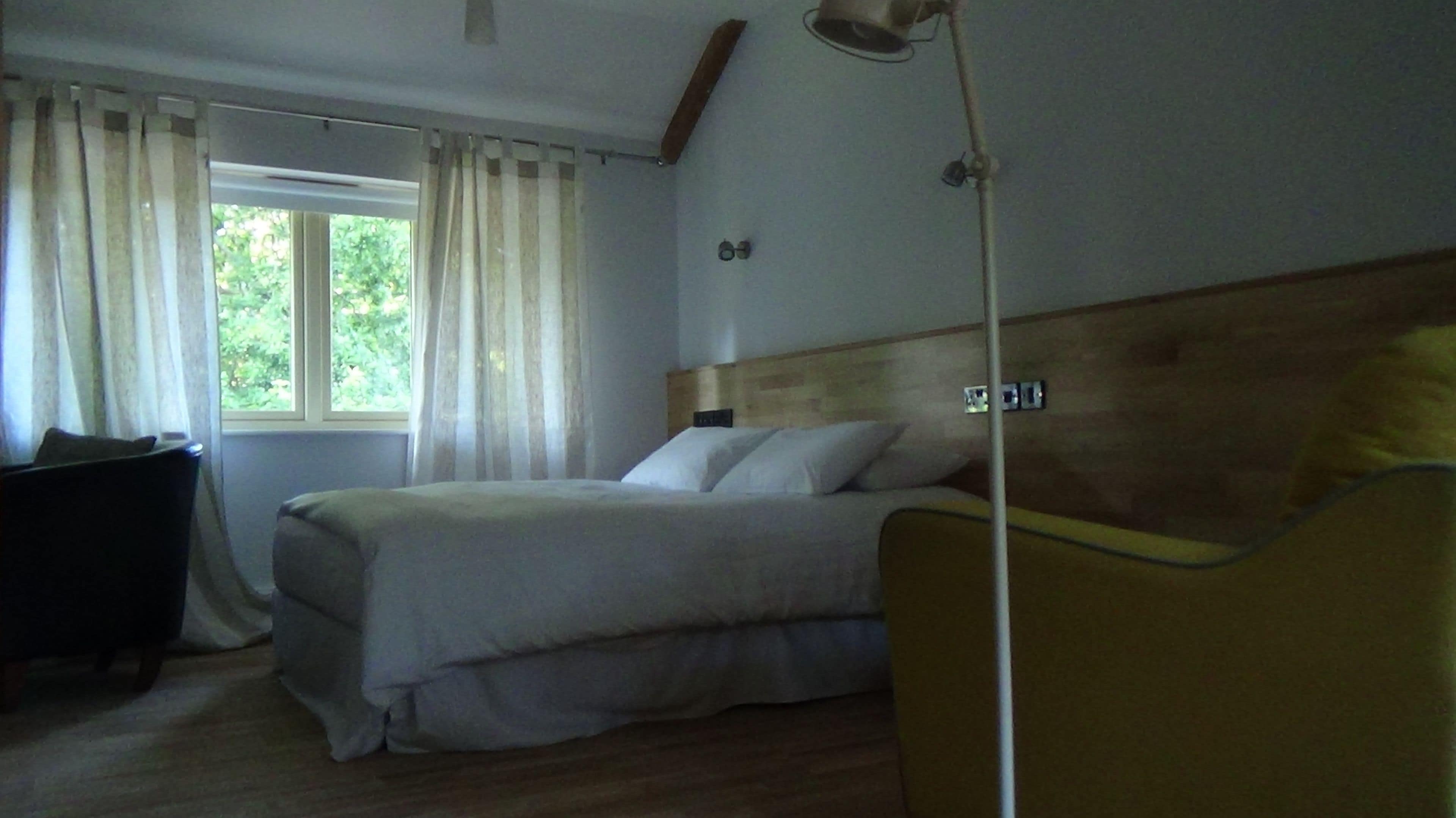 Quarto com cama, mesa de cabeceira, abajur, janela com cortinas e vista para o verde lá fora.