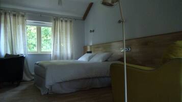 Quarto com cama, mesa de cabeceira, abajur, janela com cortinas e vista para o verde lá fora.