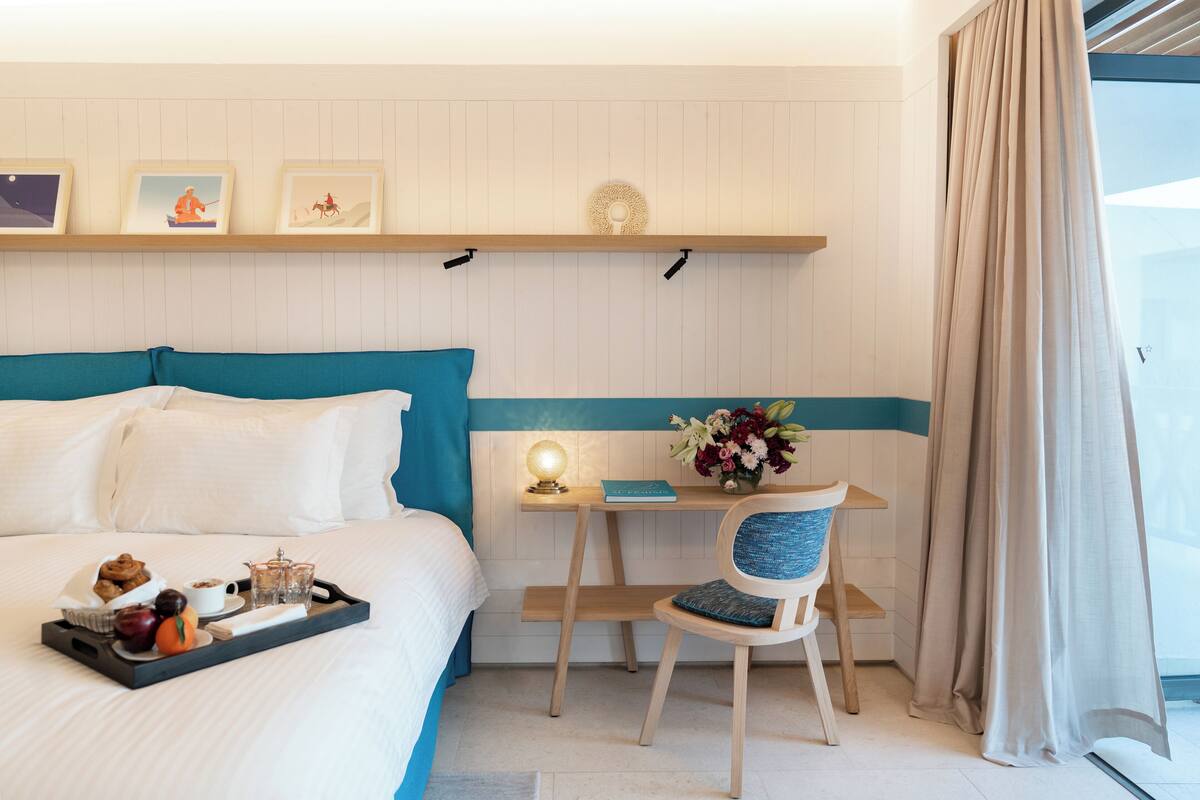Quarto de hotel com uma cama, uma escrivaninha com cadeira, uma bandeja com itens de café da manhã e uma janela com cortinas.