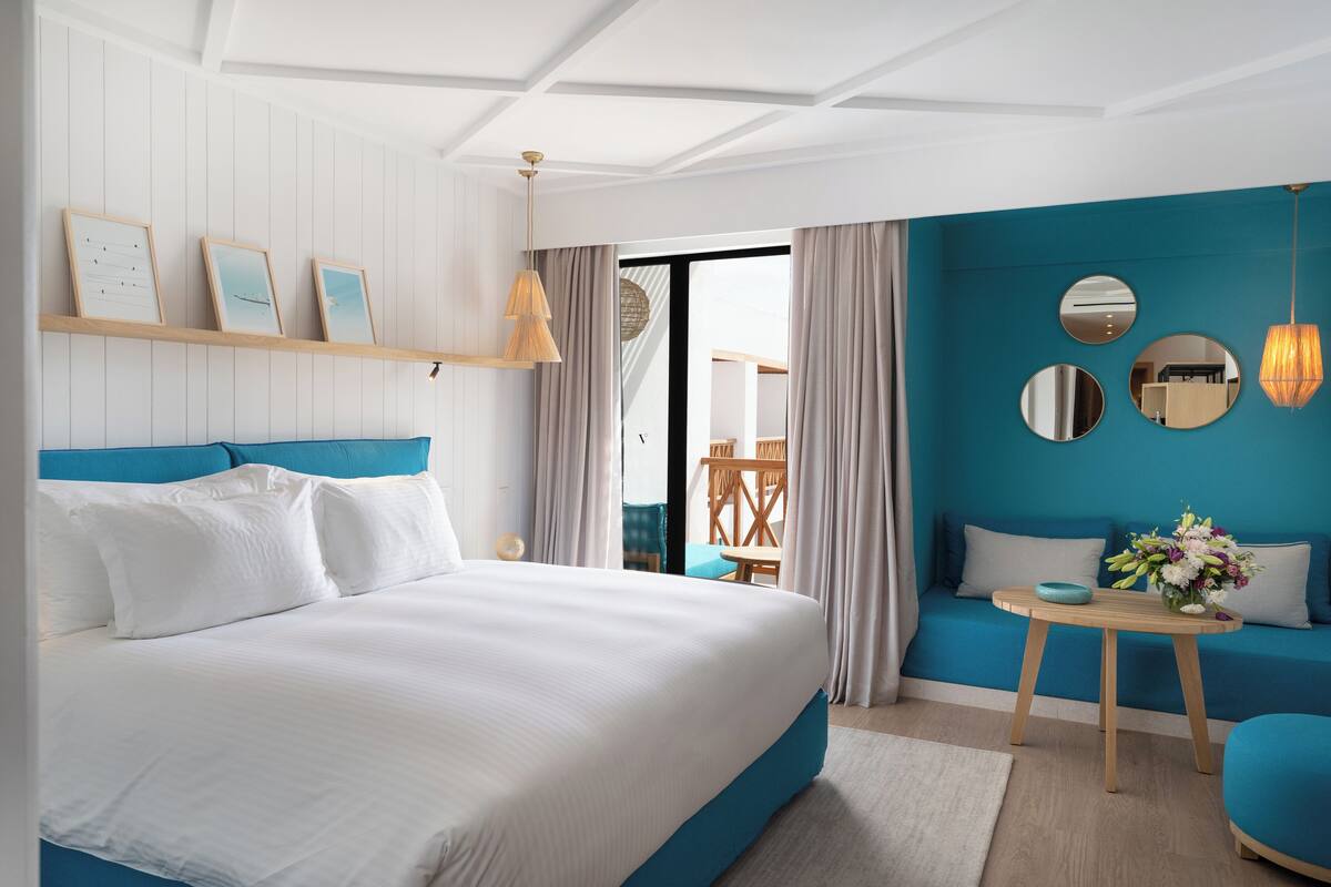 Quarto de hotel moderno com uma cama grande, um sofá azul e uma mesa pequena com um vaso de flores.