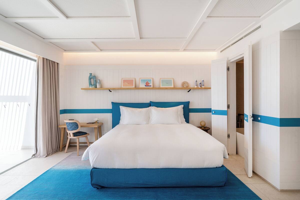 Quarto de hotel com uma cama grande, uma escrivaninha e uma cadeira. Há uma estante com objetos decorativos sobre a cama.