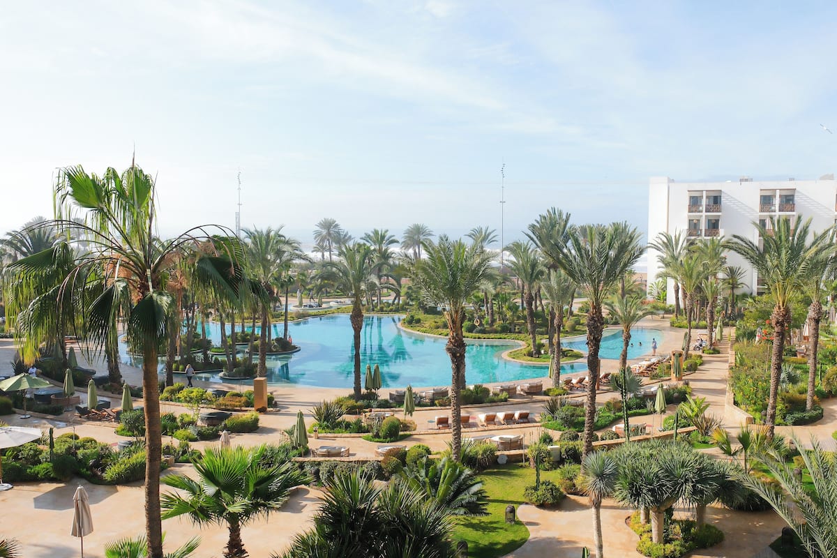 Resort com várias piscinas rodeadas por palmeiras e vegetação, um edifício grande com varandas, e céu limpo.