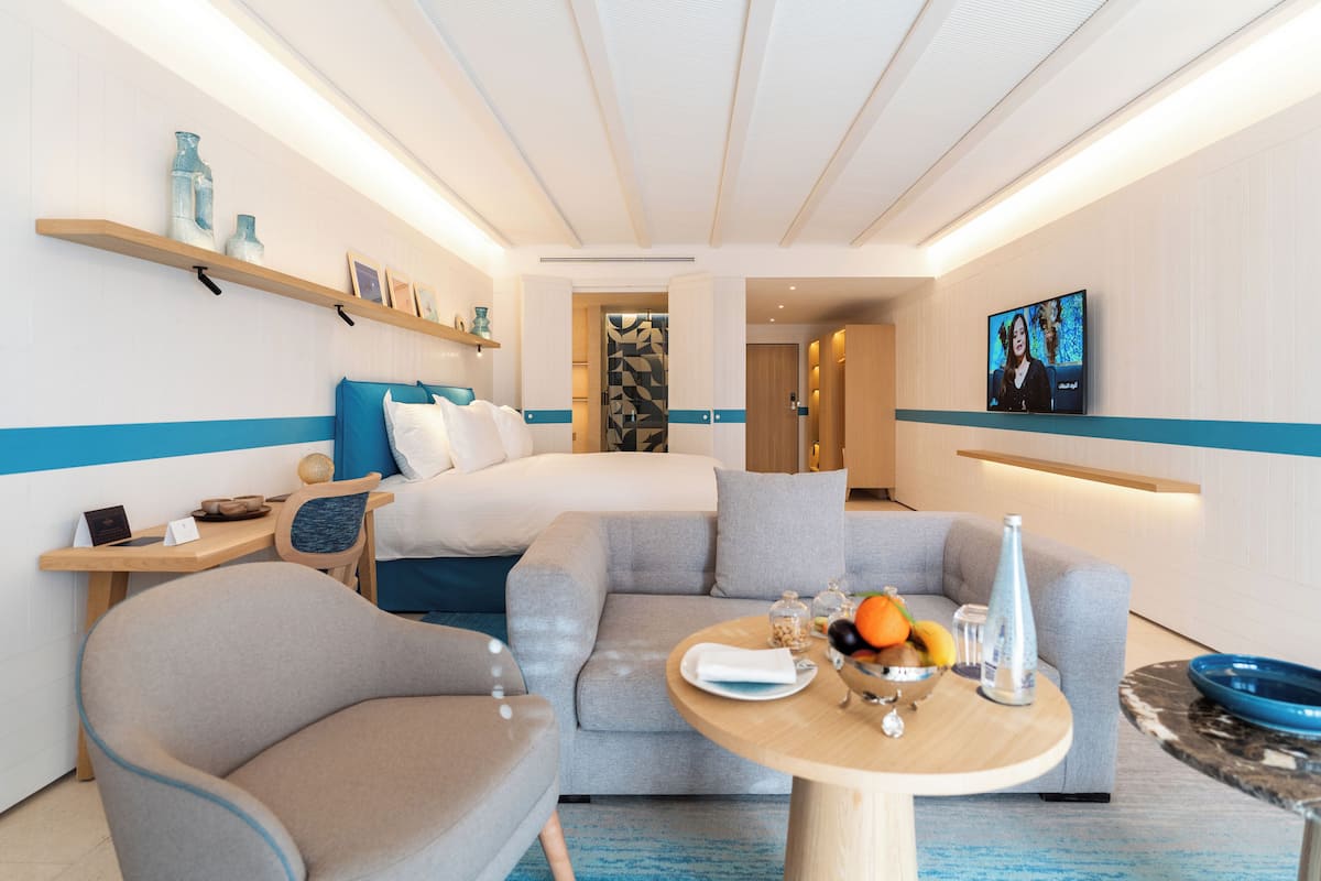 Quarto de hotel moderno com cama, sofá e área de refeições com mesa e cadeiras.