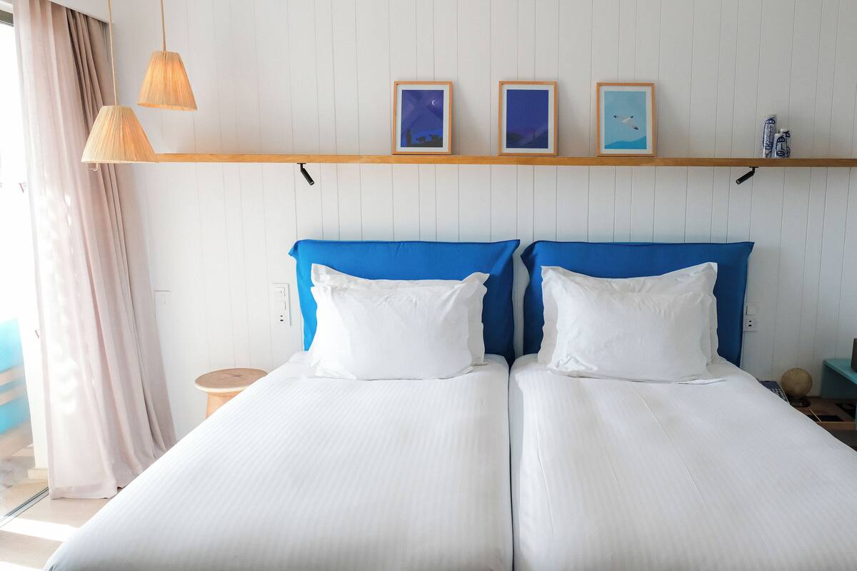 Uma cama de casal com lençóis brancos e cabeceiras azuis, uma prateleira de madeira com quadros e uma mesa de cabeceira com controle remoto e caixa de lenços.