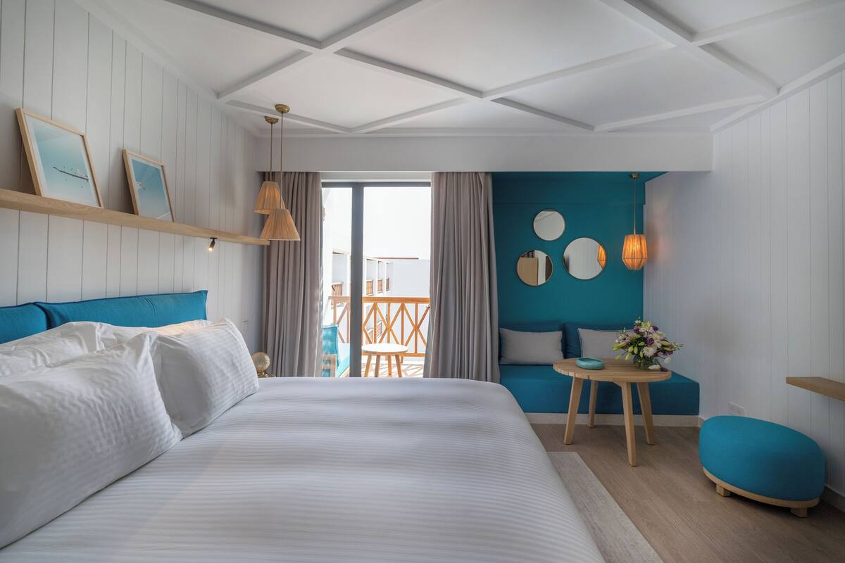 Quarto de hotel com uma cama grande, área de estar com uma mesa e varanda com guarda-corpo de madeira.