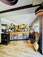 Uma área de bar com piso de madeira, banquetas, um vaso grande com plantas secas e um relógio na parede.