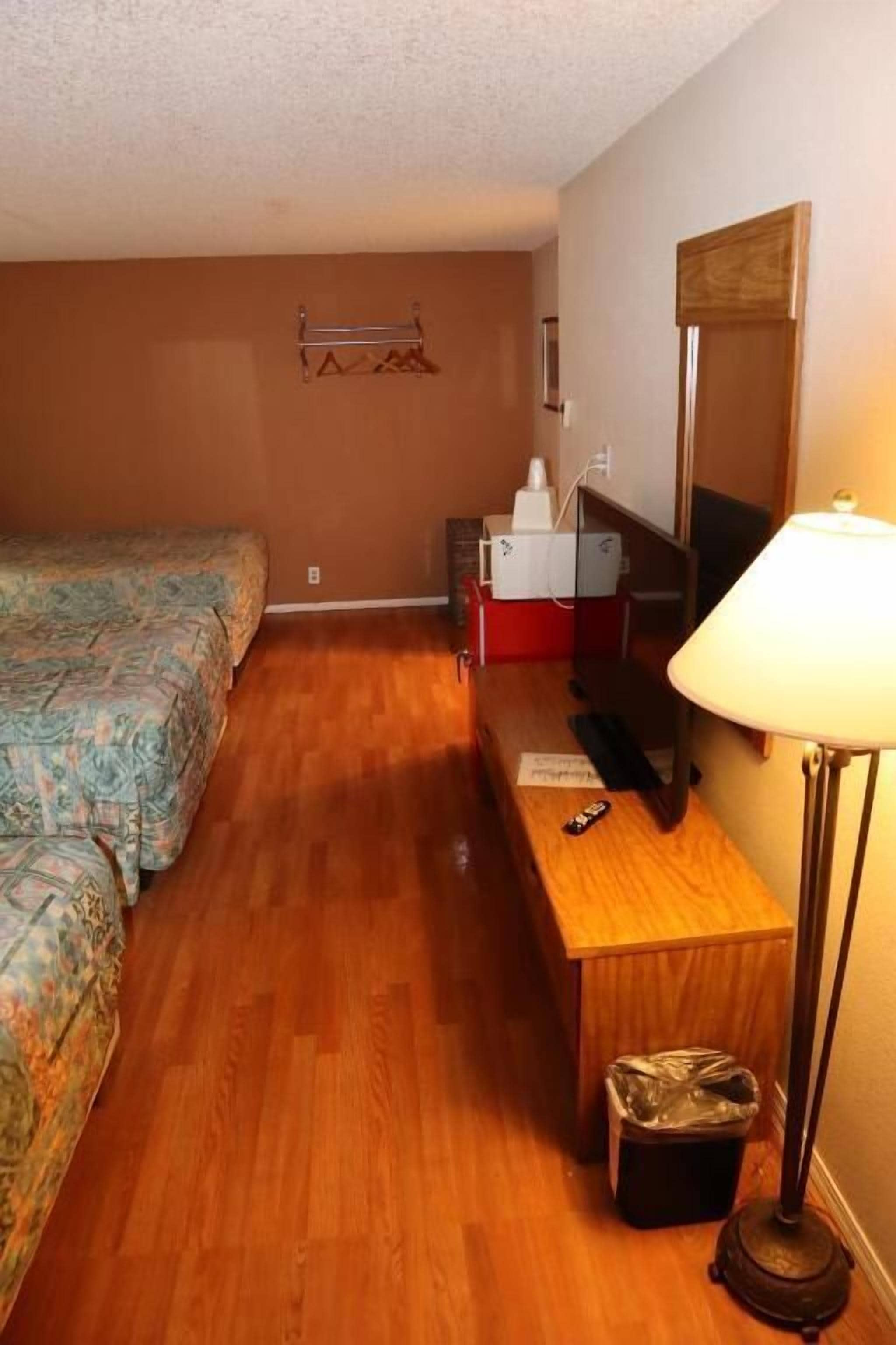 Quarto pequeno e mobiliado, com piso de madeira, uma escrivaninha, uma luminária, uma lixeira e uma cama com roupa de cama estampada.