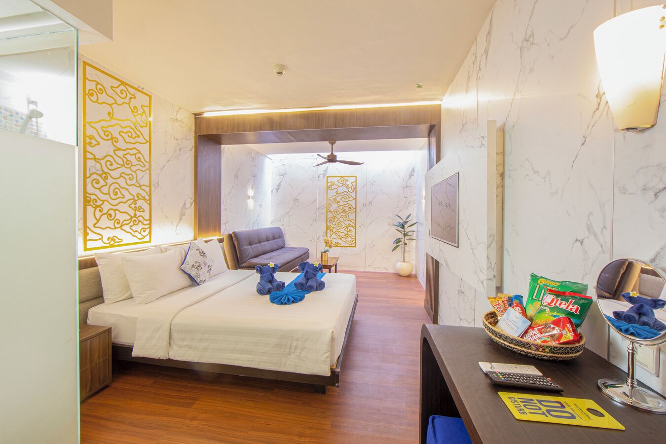 Quarto de hotel com cama, área de estar com sofá e poltronas, mesa de jantar com petiscos, espelho e ventilador de teto.