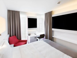 Quarto de hotel com cama, escrivaninha, cadeira e duas janelas grandes com cortinas.