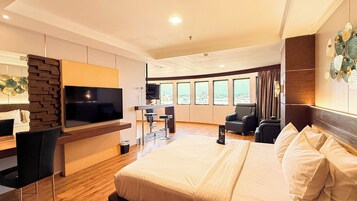Quarto de hotel moderno com uma cama grande, televisão, escrivaninha com cadeira e vista da cidade através de amplas janelas.